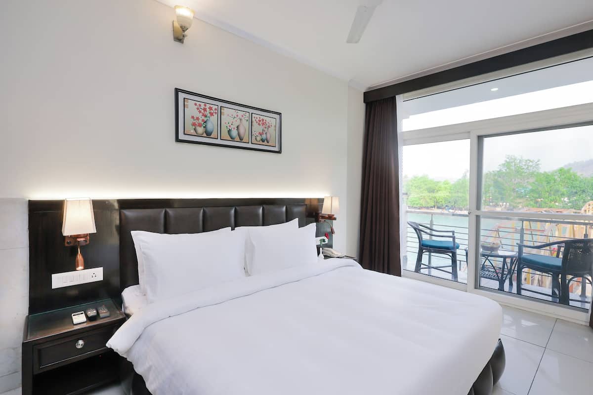 Quarto de hotel com uma cama grande, mesa de cabeceira com abajur, quadro na parede acima da cama e varanda com cadeiras e mesa.