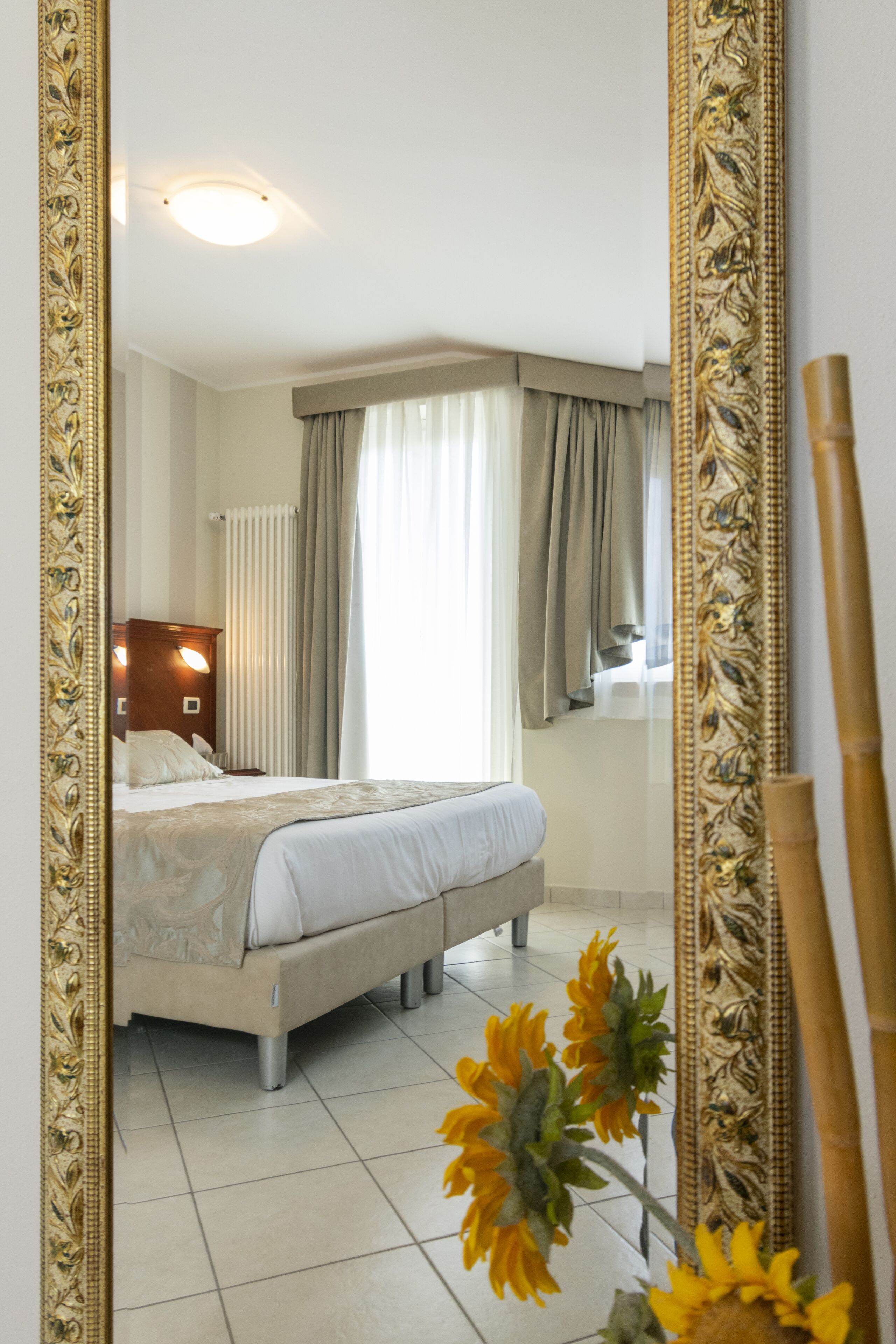 Quarto de hotel com uma cama, cortinas, uma janela e um espelho com moldura decorativa.