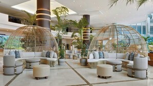 Um hall amplo com design moderno, destacando uma área de estar central com sofás e pufes, cercada por espaços abertos e luz natural.