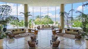 Um hall amplo com área de estar espaçosa, balcão de recepção e grandes janelas com vista para o oceano e palmeiras.