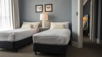 Quarto de hotel com duas camas, uma mesa de cabeceira com abajur e uma porta que dá para outro cômodo.