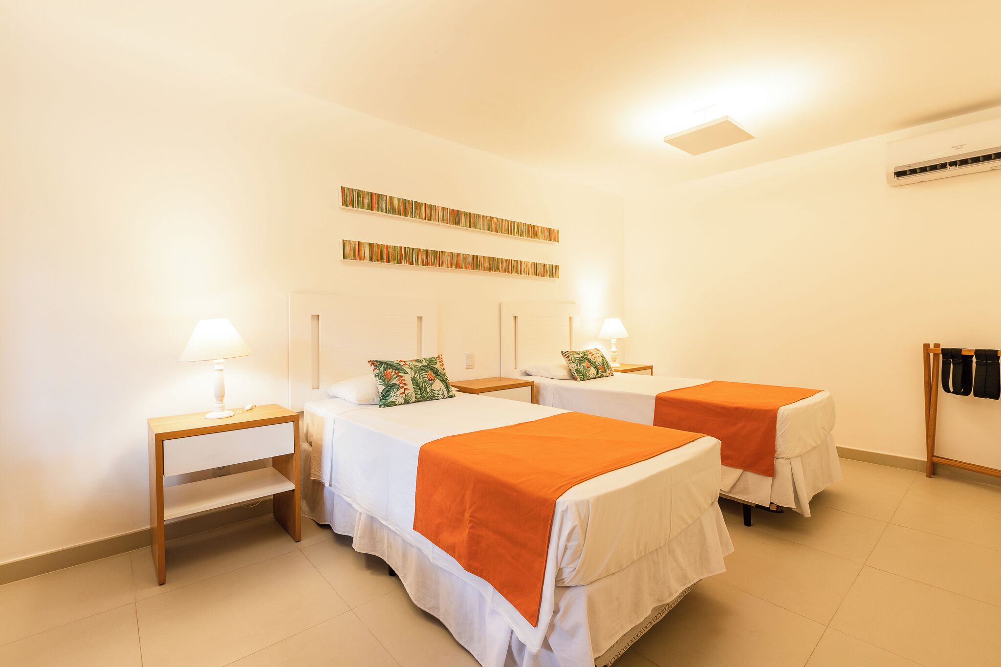 Quarto de hotel com duas camas, cada uma com manta laranja, roupa de cama branca e mesa de cabeceira com abajur. Há unidades de ar condicionado fixadas na parede e uma estante com objetos decorativos.
