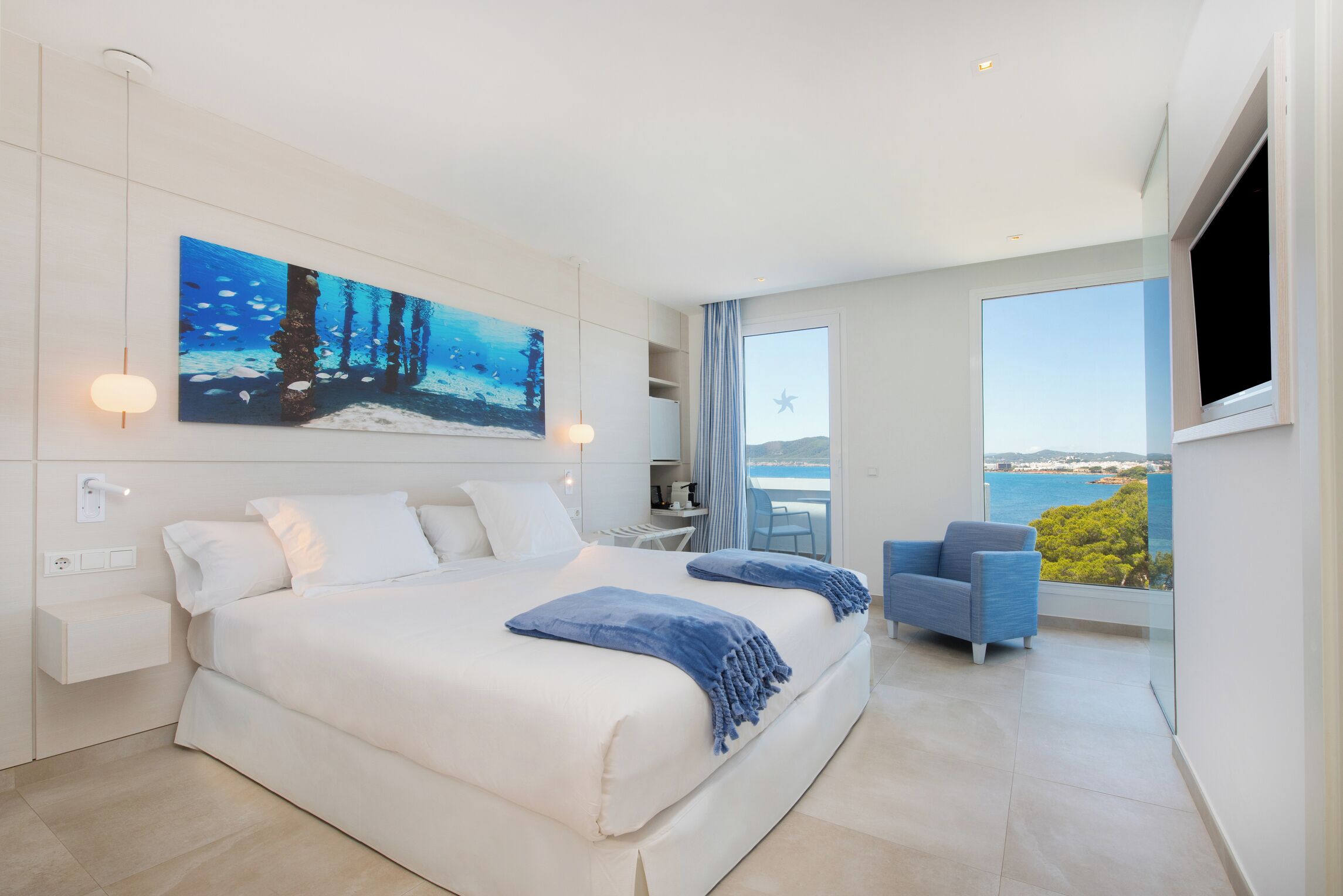 Quarto moderno com uma cama grande, uma pintura de uma cena subaquática, uma varanda com vista para o mar e uma cadeira azul.