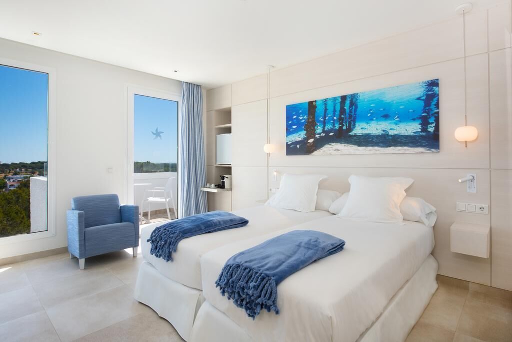 Quarto moderno com duas camas, uma janela ampla com vista, uma cadeira azul e um quadro na parede.