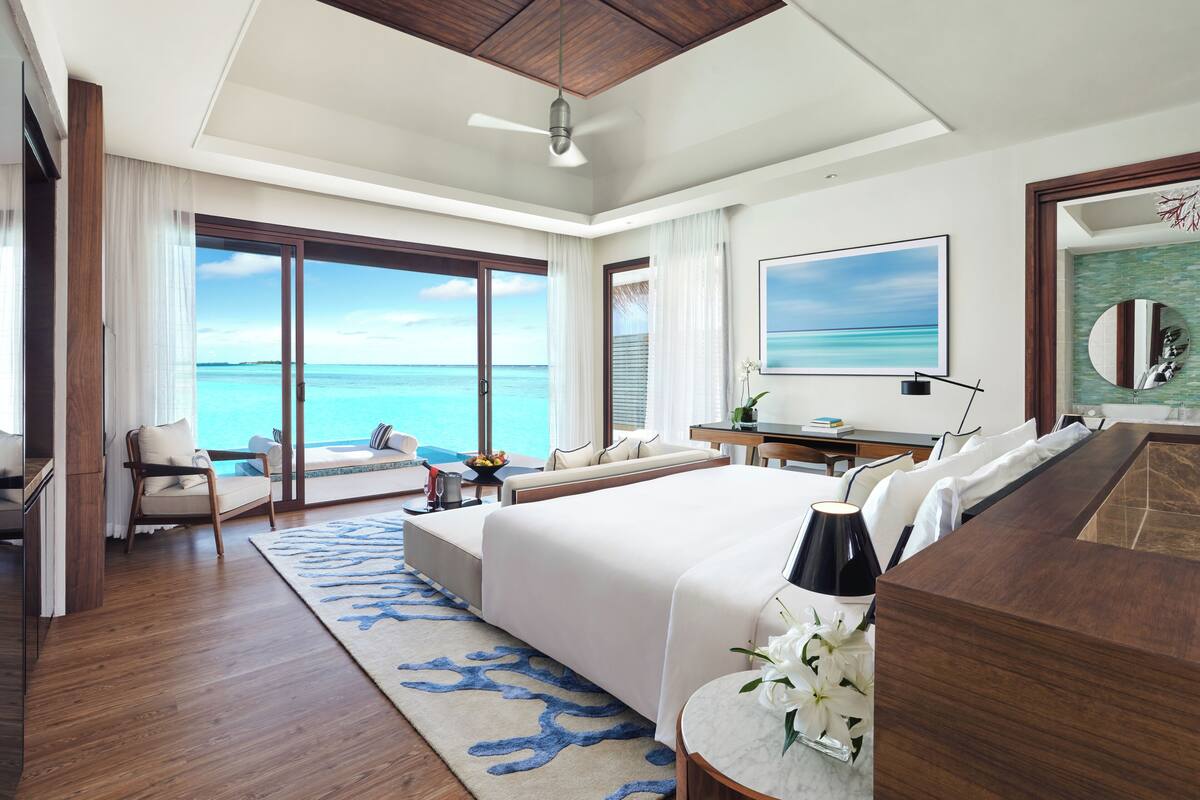 Quarto de hotel moderno com uma cama grande, uma escrivaninha, uma cadeira, vista para o oceano e um ventilador de teto.