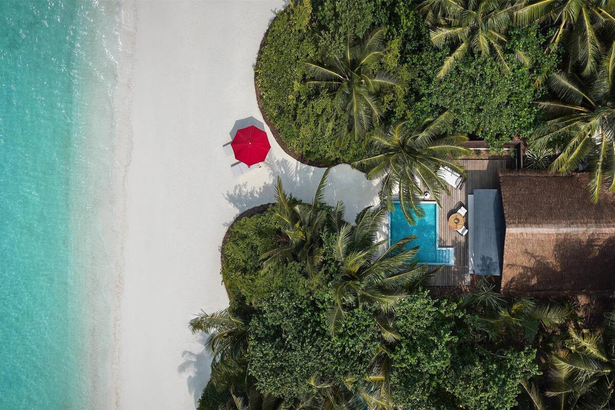 Vista aérea de uma propriedade à beira-mar, com piscina cercada por vegetação exuberante e um guarda-sol vermelho.