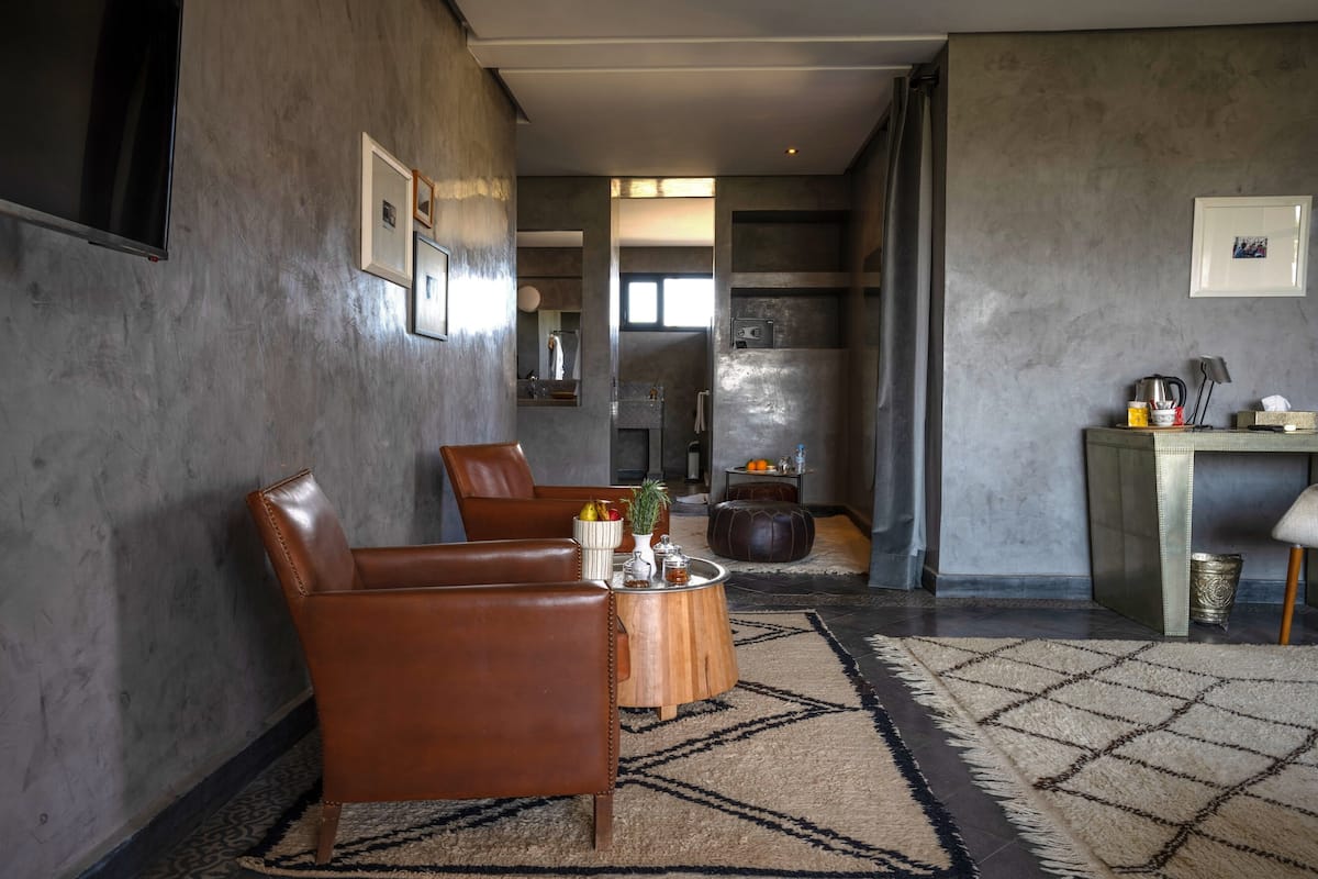 Uma sala de estar moderna com sofás de couro, uma mesa de centro com objetos decorativos, um tapete estampado e uma área de cozinha ao fundo.