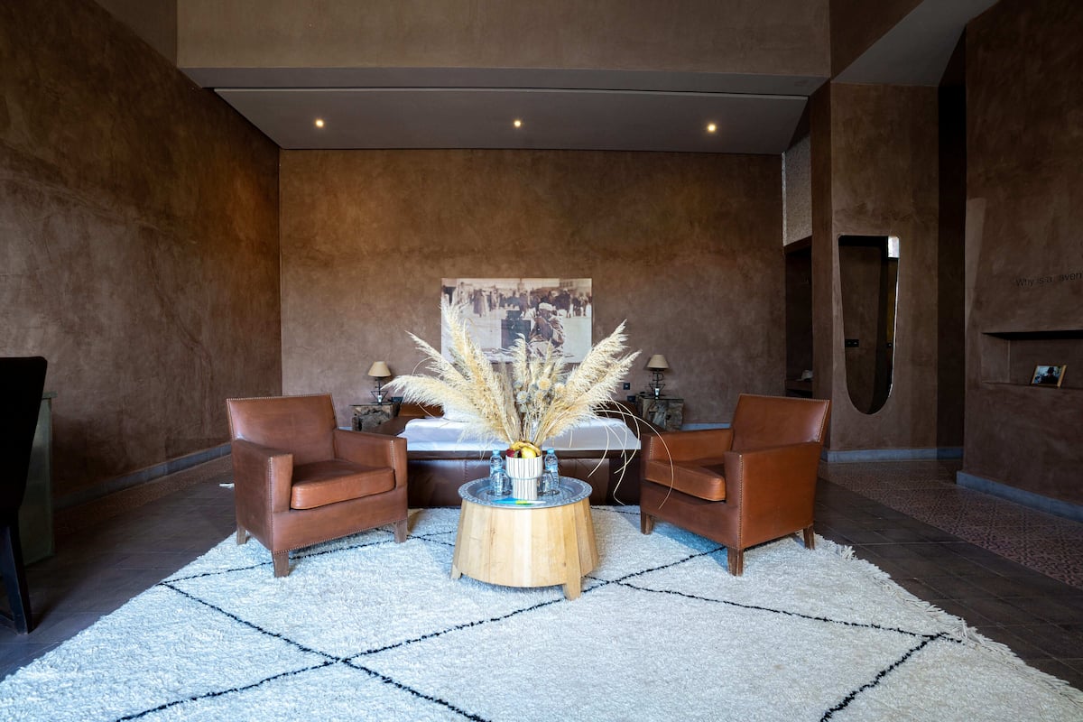 Um lobby com duas poltronas de couro, uma mesa de centro redonda de madeira com um vaso de plantas secas e uma grande fotografia emoldurada na parede.