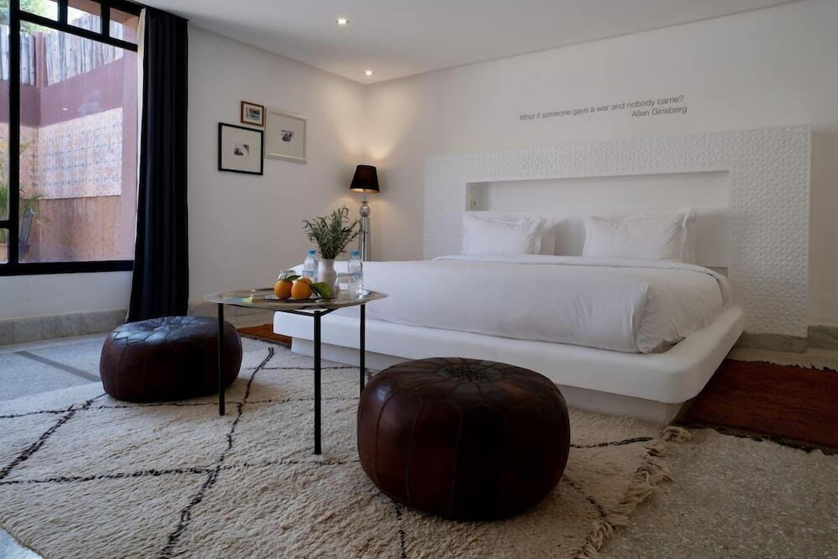 Quarto moderno com uma cama grande, uma mesa redonda com abajur e dois pufes.