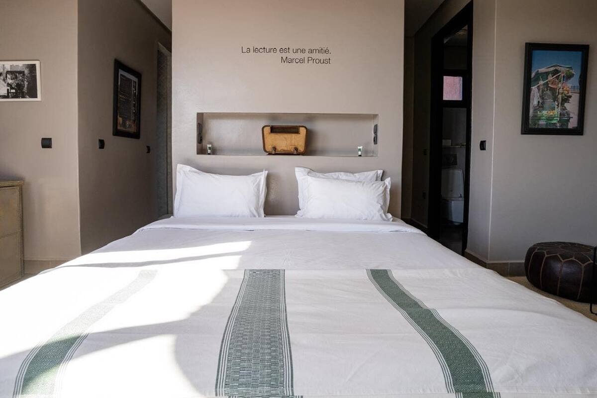 Quarto de hotel com uma cama grande, cabeceira de madeira e uma frase na parede.