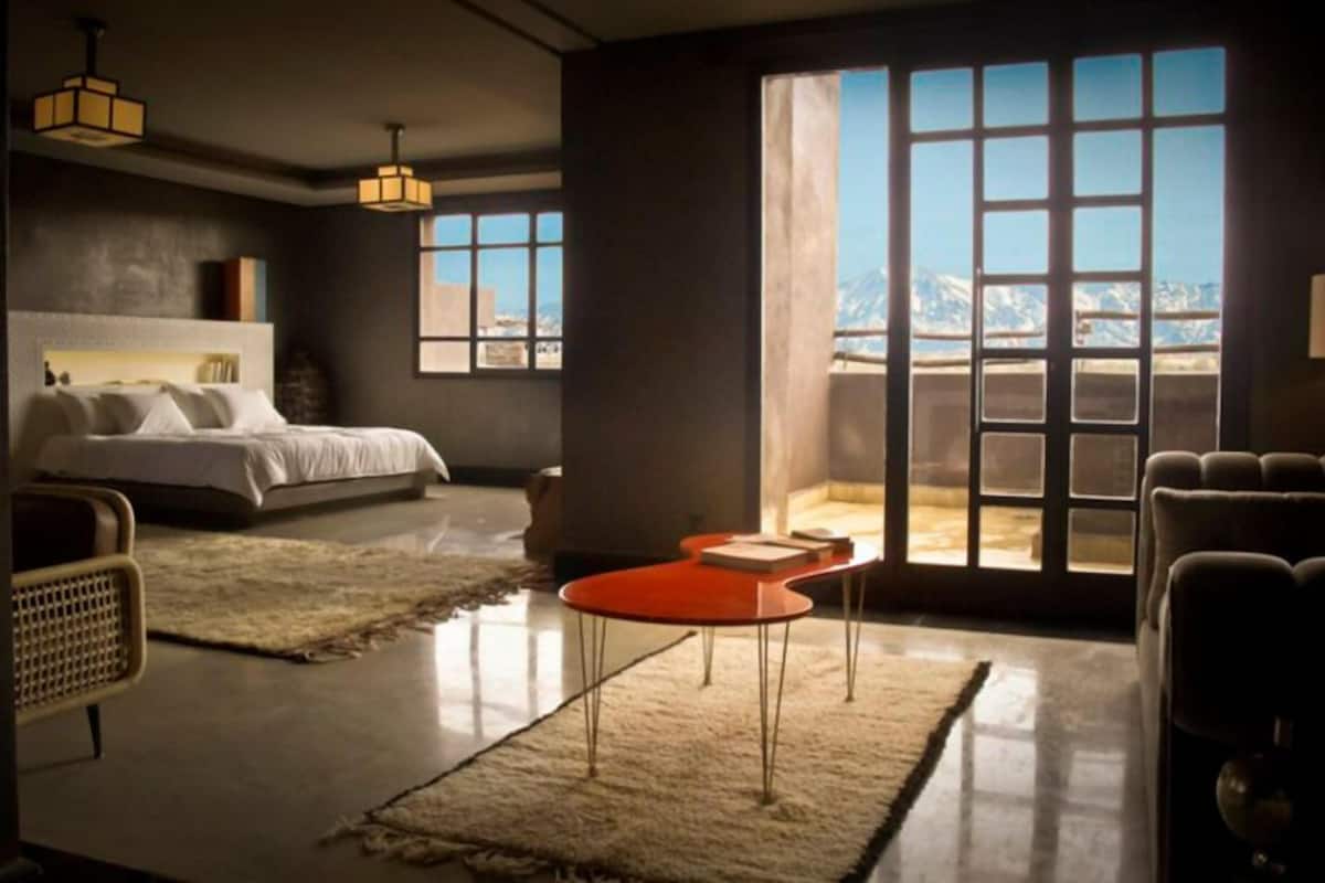 Quarto moderno com uma cama grande, mesa de cabeceira vermelha e vista para uma paisagem nevada através de uma janela ampla.