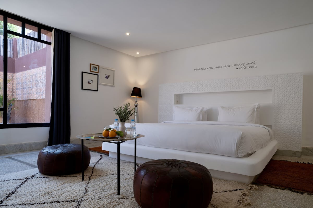 Quarto moderno com cama grande, mesa de cabeceira com frutas e garrafas de água, abajur e uma frase decorativa na parede.