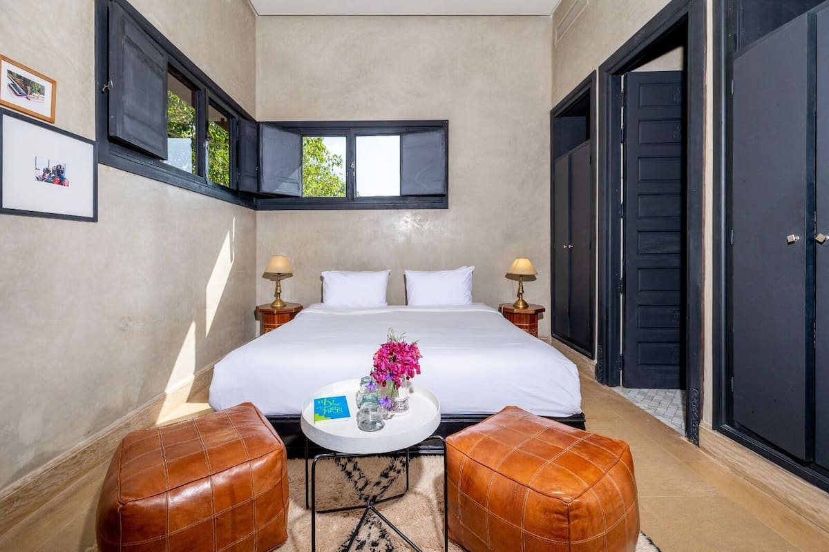 Quarto com uma cama grande, duas mesas de cabeceira com luminárias, uma mesinha redonda com um vaso de flores e dois pufes de couro.