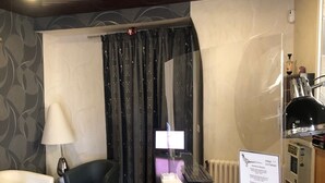 Uma área de recepção de hotel com um balcão curvo, uma cadeira branca, um poltrona escura, um abajur e uma janela com cortinas.