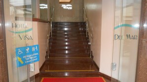 Entrada de hotel com uma escadaria que leva ao andar superior. Há uma placa indicando uma entrada lateral para hóspedes com deficiência. O nome do hotel 'VINA DEL MAR' está visível nas paredes e no carpete.