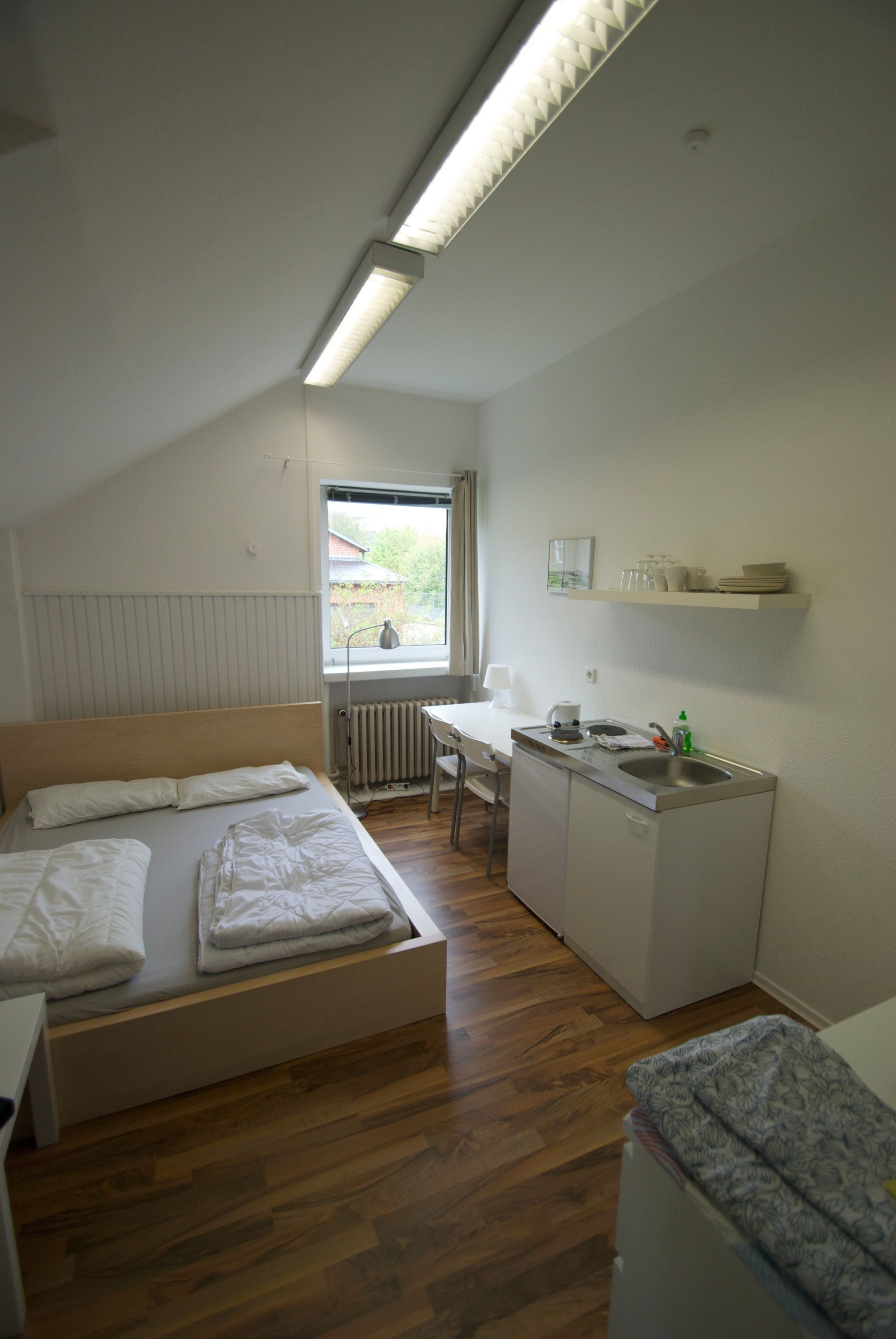 Quarto pequeno e bem iluminado com uma cama, uma escrivaninha, uma cozinha compacta e uma janela com vista para áreas verdes.
