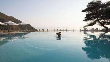 Uma piscina com espreguiçadeiras e guarda-sóis, cercada por uma vedação e com montanhas ao fundo.
