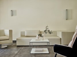 Uma sala de estar minimalista com um sofá branco, uma poltrona preta, uma mesa de centro de vidro com livros e um vaso com planta.