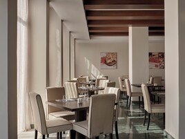 Uma sala de jantar moderna com pé direito alto e iluminação natural, com mesas e cadeiras dispostas para os hóspedes.