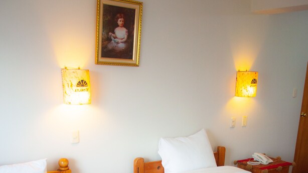 Quarto de hotel com duas camas, cada uma com lençóis brancos e um cobertor listrado colorido. Há luminárias fixadas na parede e um quadro emoldurado.