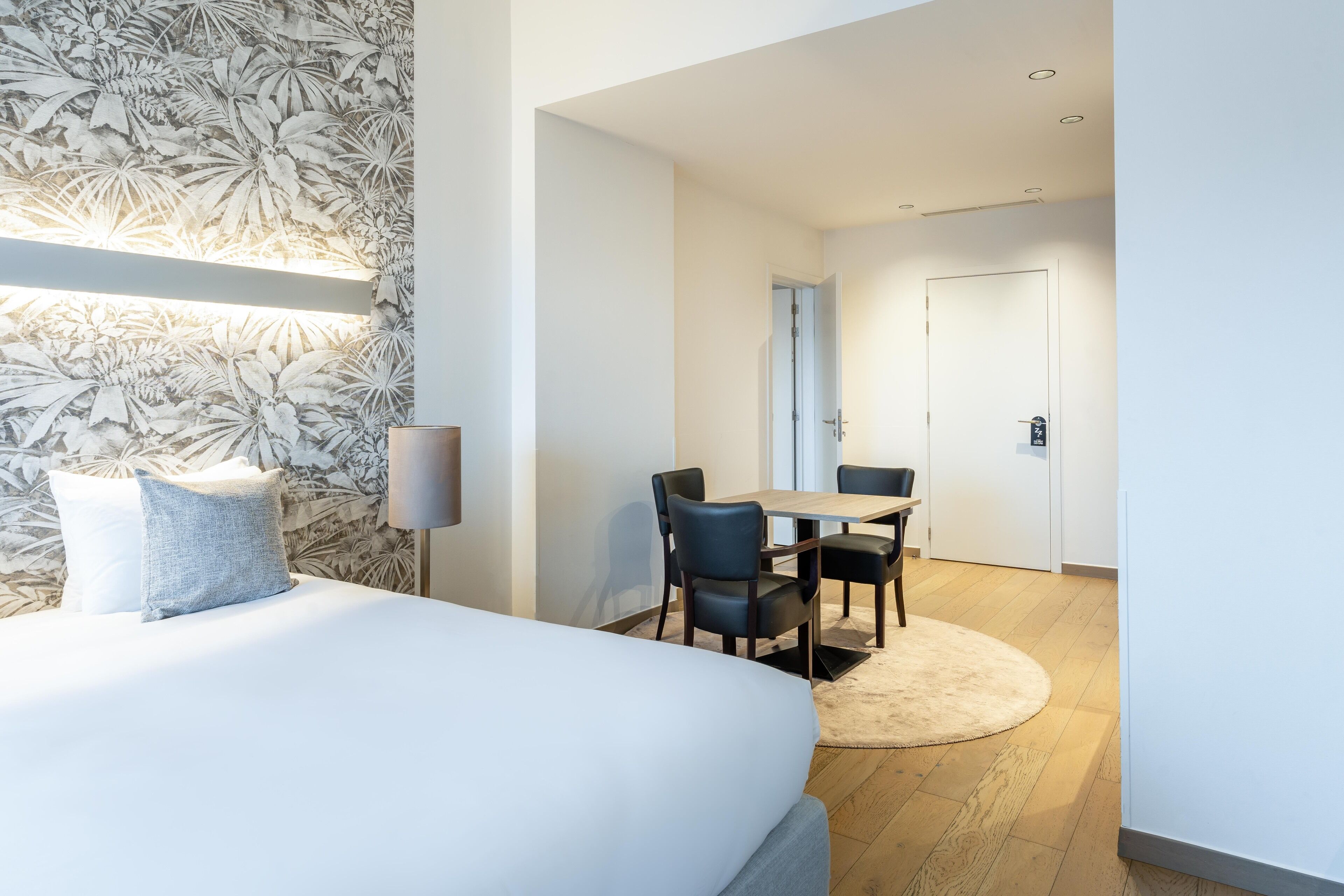 Quarto de hotel com uma cama, uma mesa e uma cadeira. Uma porta ao fundo.