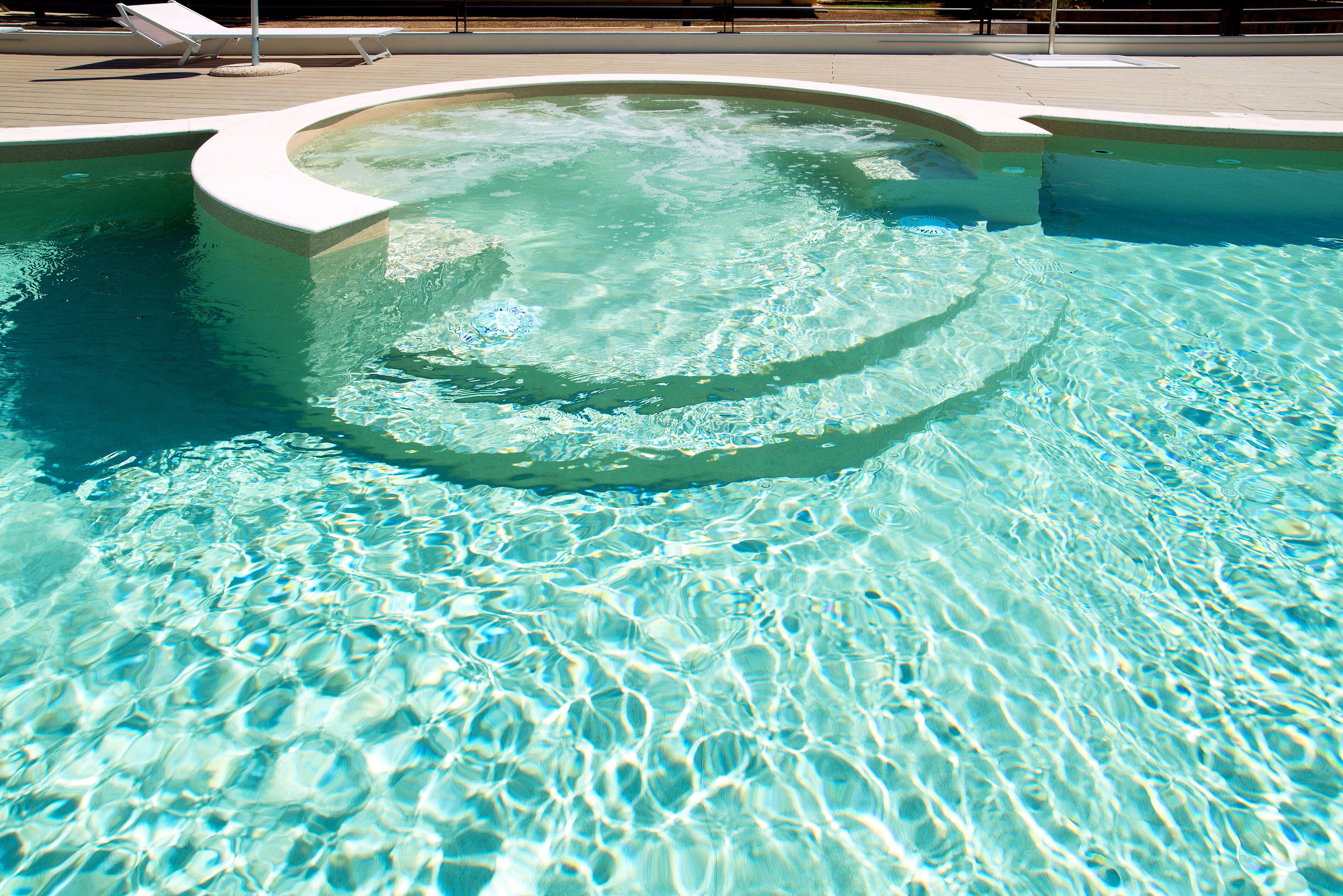 Piscina transparente com borda curva e área de deck revestida com espreguiçadeiras.