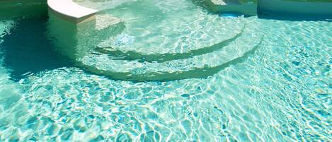 Piscina transparente com borda curva e área de deck revestida com espreguiçadeiras.
