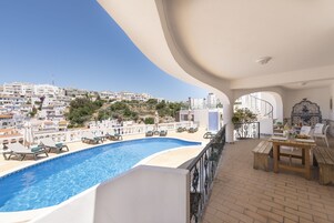 Uma piscina no terraço com espreguiçadeiras e área de refeições, com vista para um bairro residencial.
