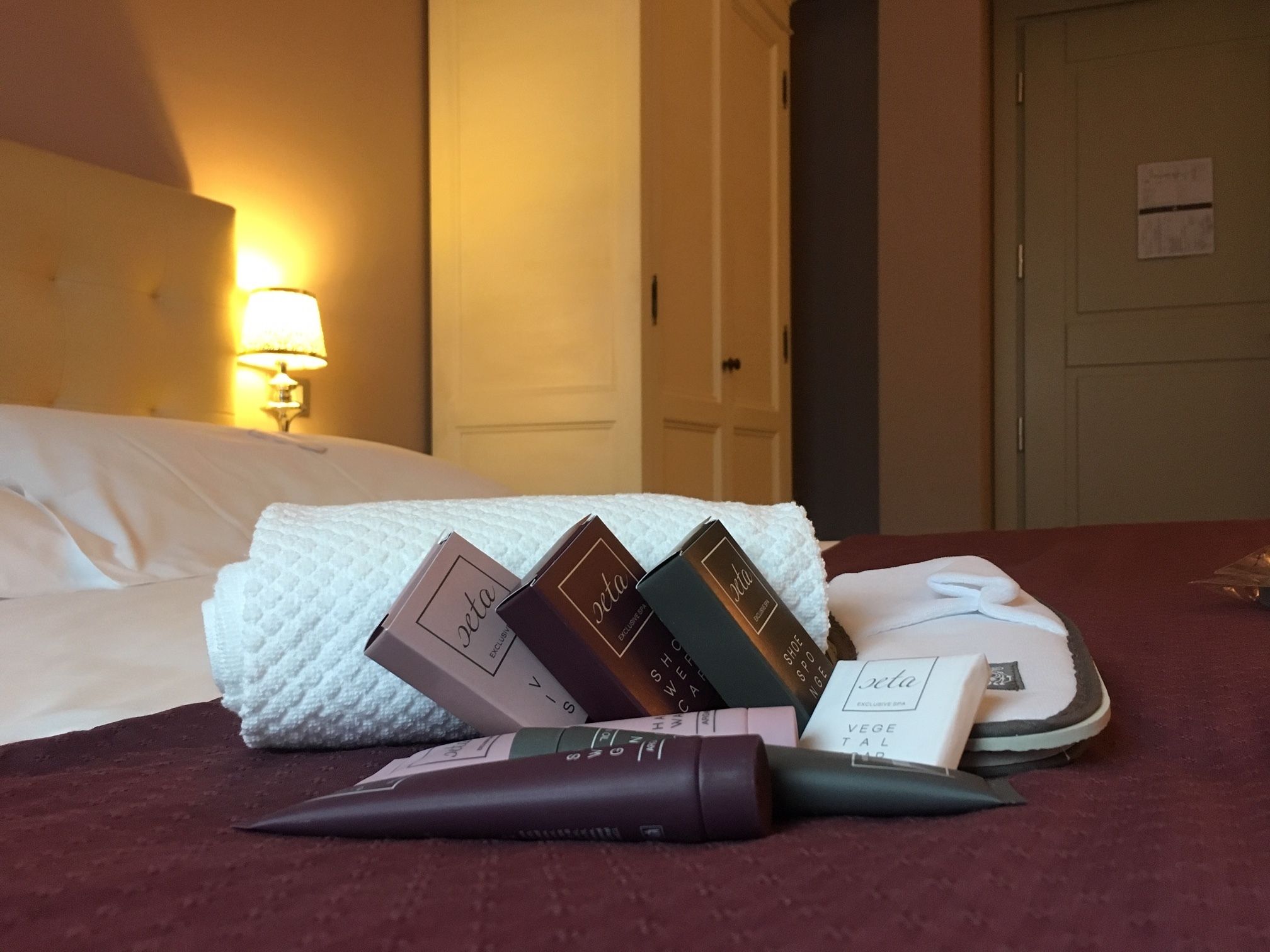 Cama bem arrumada com travesseiro e toalha brancos, um conjunto de produtos de higiene pessoal incluindo escova de dentes, lâmina de barbear e um tubo de creme, e uma luminária na mesa de cabeceira.