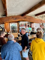 Um espaço de bar rústico com vigas de madeira, parede de pedra e diversos copos e garrafas em prateleiras.