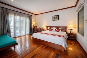 Quarto de hotel com piso de madeira, cama com roupa de cama branca e almofadas vermelhas e brancas, escrivaninha com cadeira, mesa de cabeceira com abajur e janela com cortinas.
