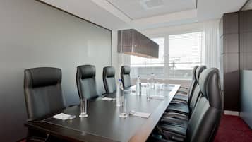 Uma sala de conferência com uma mesa comprida, cadeiras de couro preto, garrafas de água e uma grande janela com persianas.