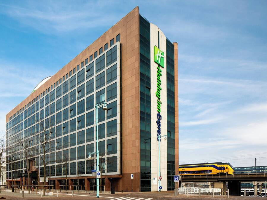 Edifício moderno de hotel com placa verde e branca em destaque, grandes janelas e faixa de pedestres.