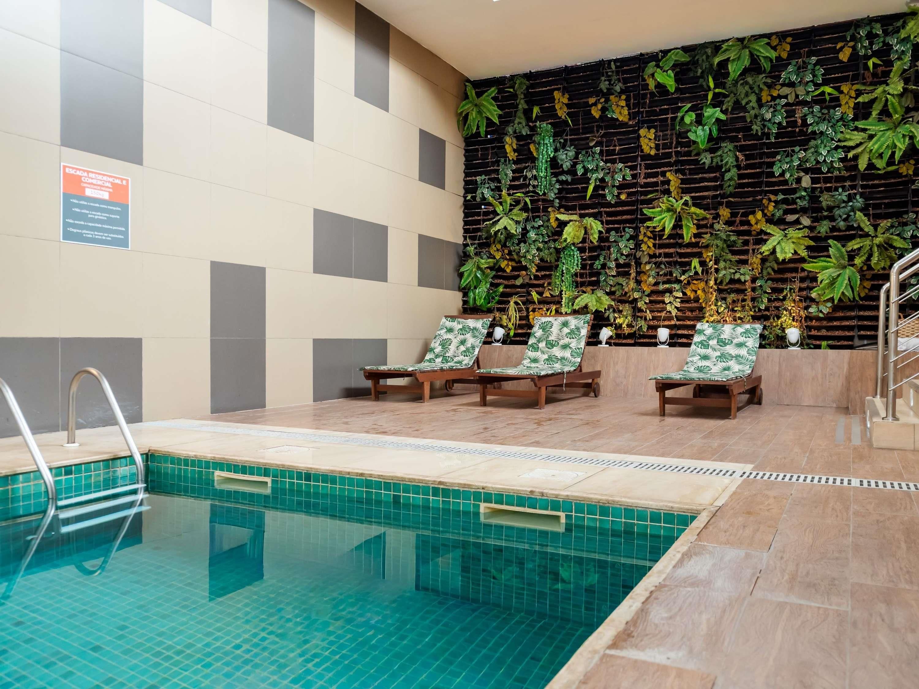 Piscina interna com chão e paredes revestidos, rodeada por cadeiras de lounge com estampas tropicais.