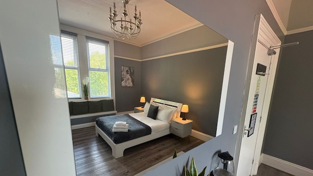 Quarto moderno com cama, abajures, um lustre, janela com persianas e uma planta em vaso.