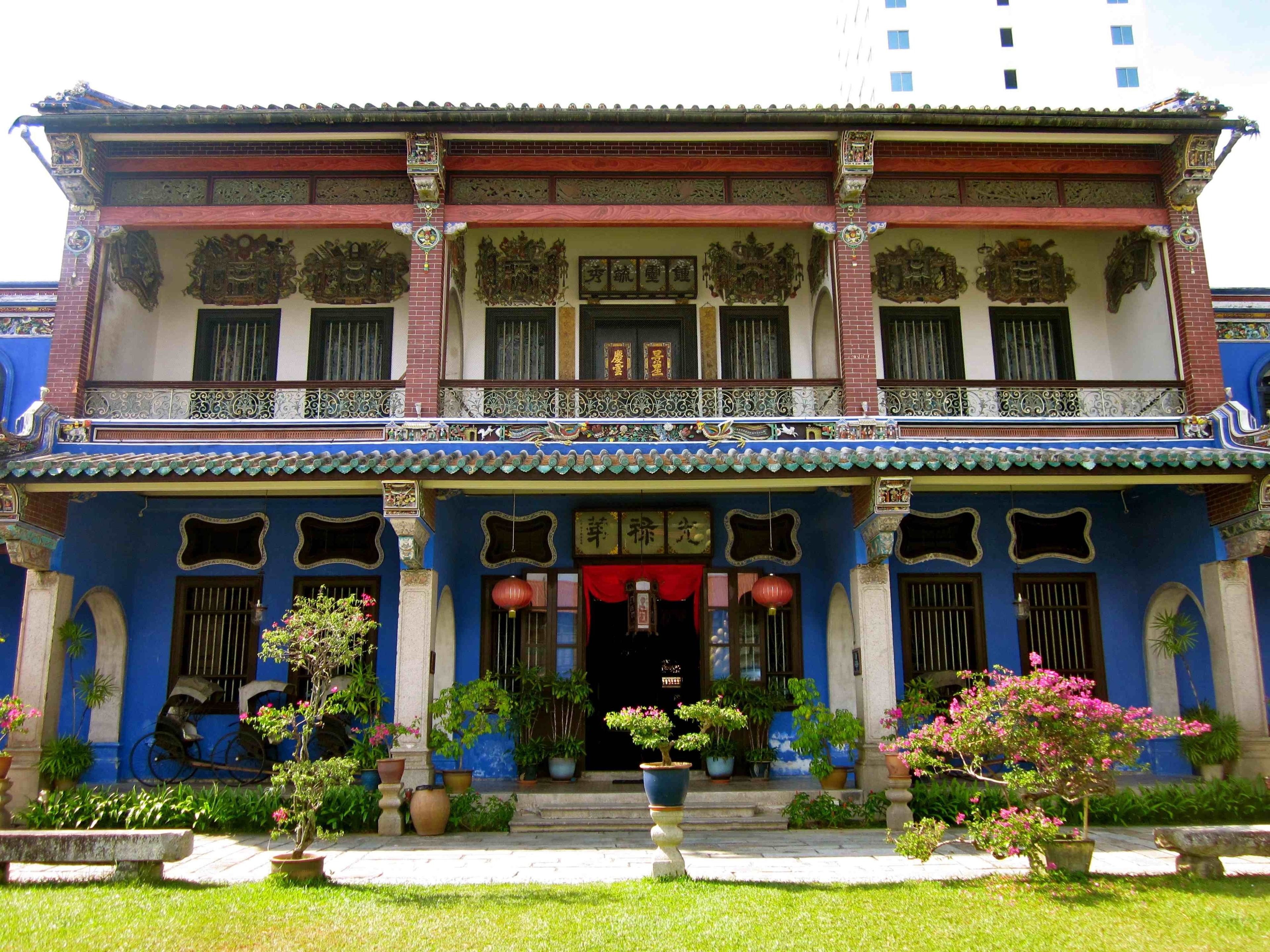 Um edifício tradicional com estrutura de dois andares, decorações elaboradas e um jardim bem cuidado com plantas e flores em vasos.