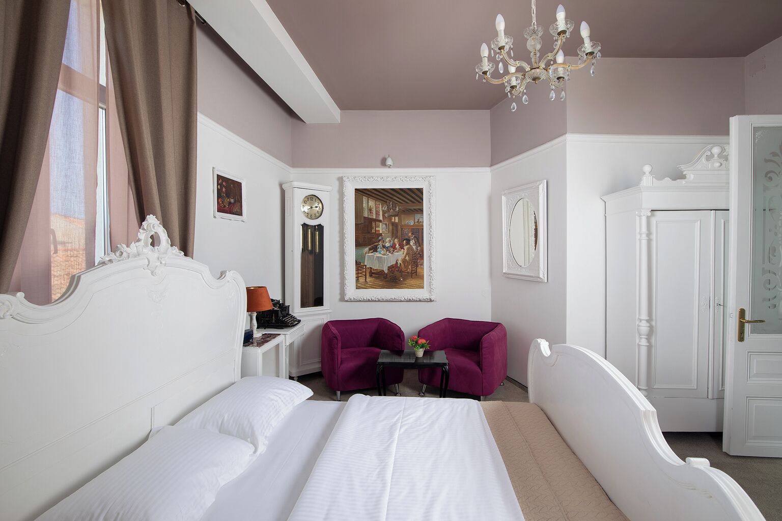 Quarto com uma cama grande, uma escrivaninha com luminária, duas cadeiras, um candelabro e um quadro na parede.