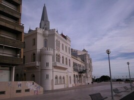 Um edifício grande e histórico com uma torre imponente e janelas em arco, situado em área urbana com praça pavimentada e bancos.