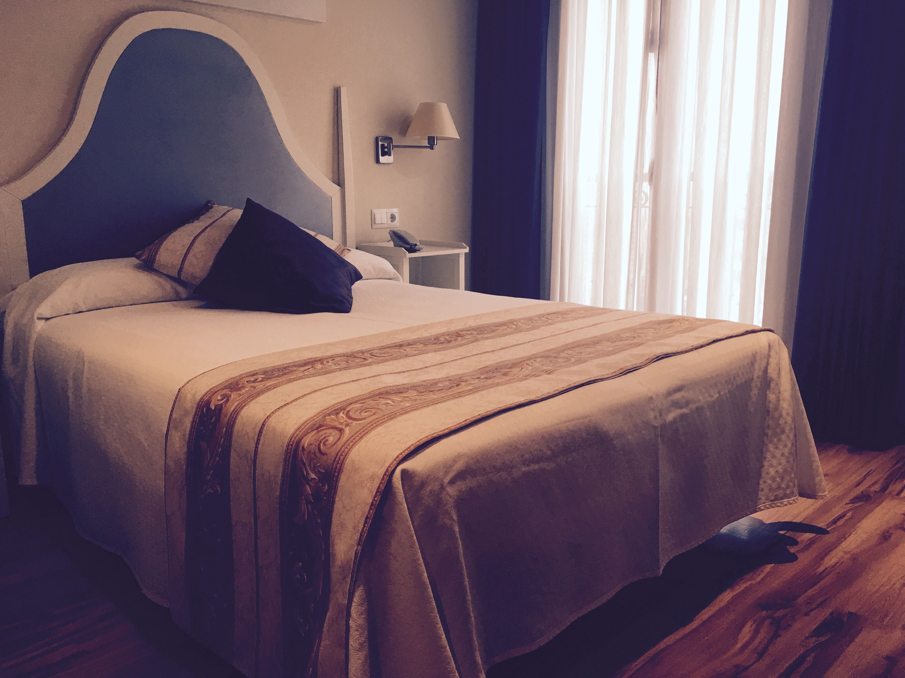 Uma cama bem arrumada com colcha estampada e almofadas, uma mesa de cabeceira com abajur e uma janela com cortinas.