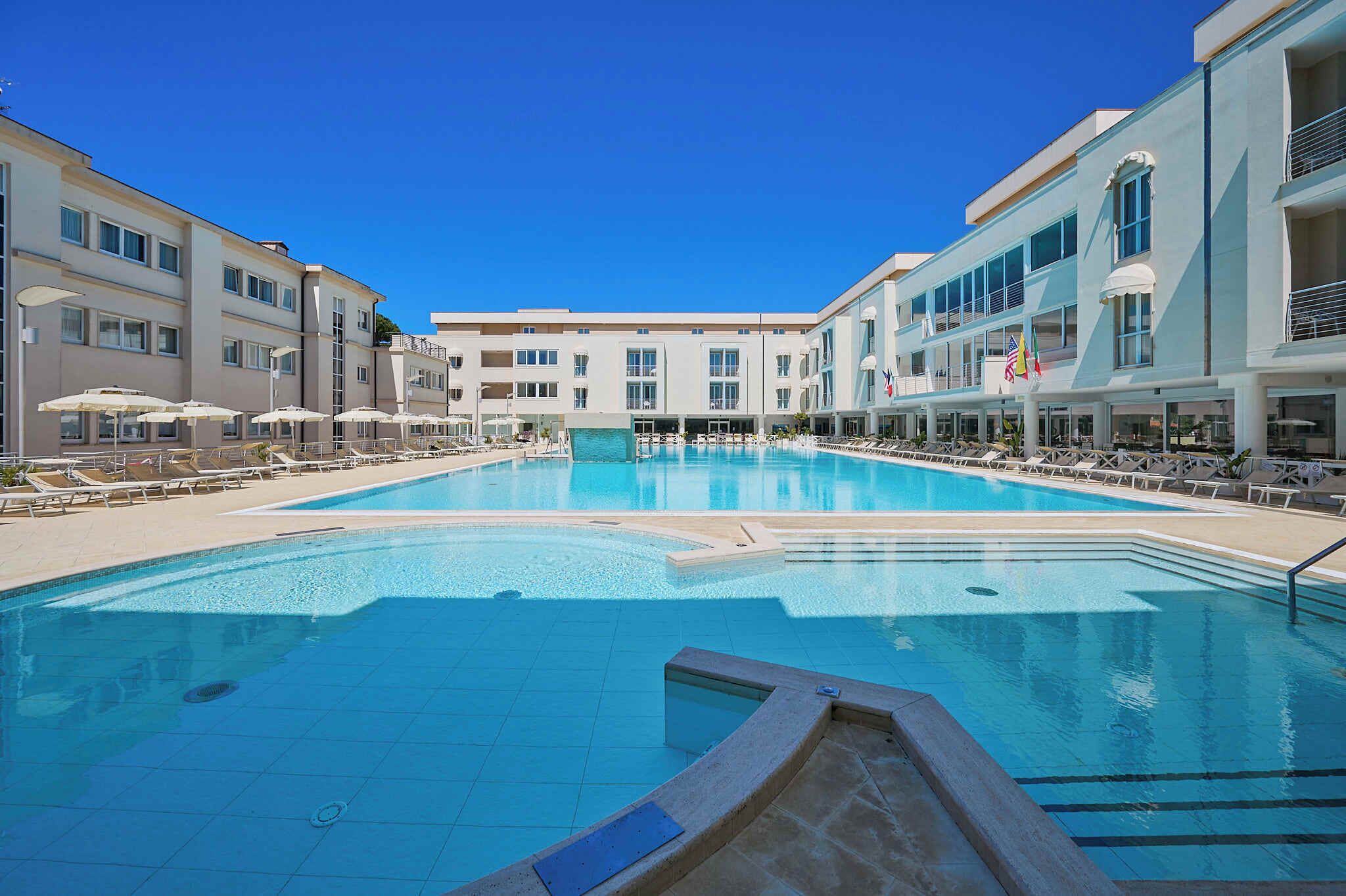 Piscina com espreguiçadeiras e guarda-sóis, rodeada por edifícios altos com varandas.