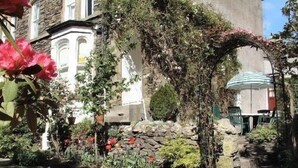 Um jardim com um caminho de pedra, uma pérgula coberta de plantas trepadoras e um edifício com fachada branca ao fundo.