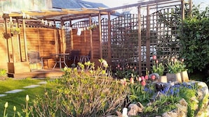 Uma pérgola de madeira com cadeiras, um muro de pedra com plantas e uma cerca de madeira com roupa pendurada.