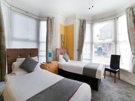 Quarto de hotel com duas camas, uma poltrona, uma mesinha de cabeceira e um quadro na parede.
