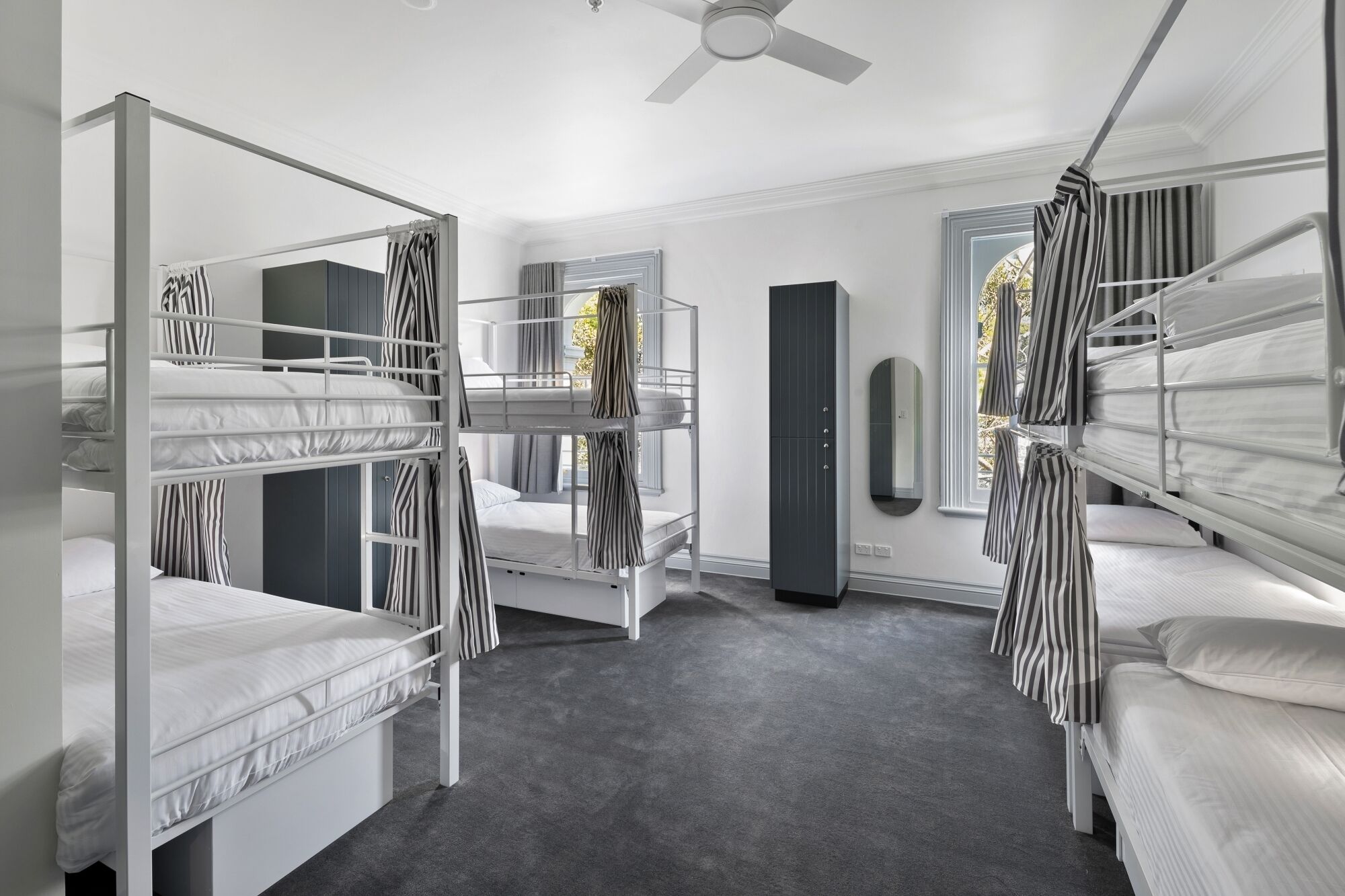 Quarto com beliches, ventoinha no teto e uma grande janela com cortinas.