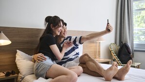 Um casal tira um selfie em uma cama em quarto de hotel, com cabeceira de madeira, abajur e janela com cortinas.