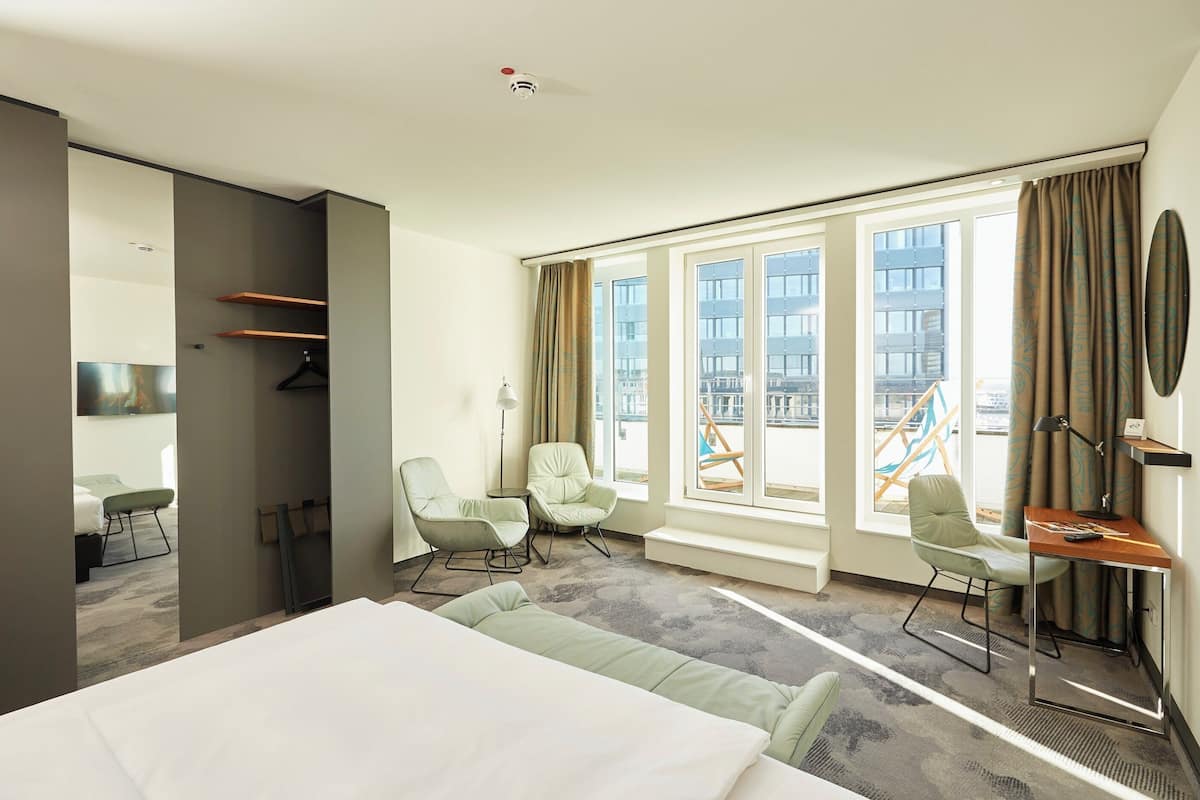 Quarto de hotel moderno com uma cama grande, duas poltronas, uma escrivaninha e vista da cidade através de amplas janelas.