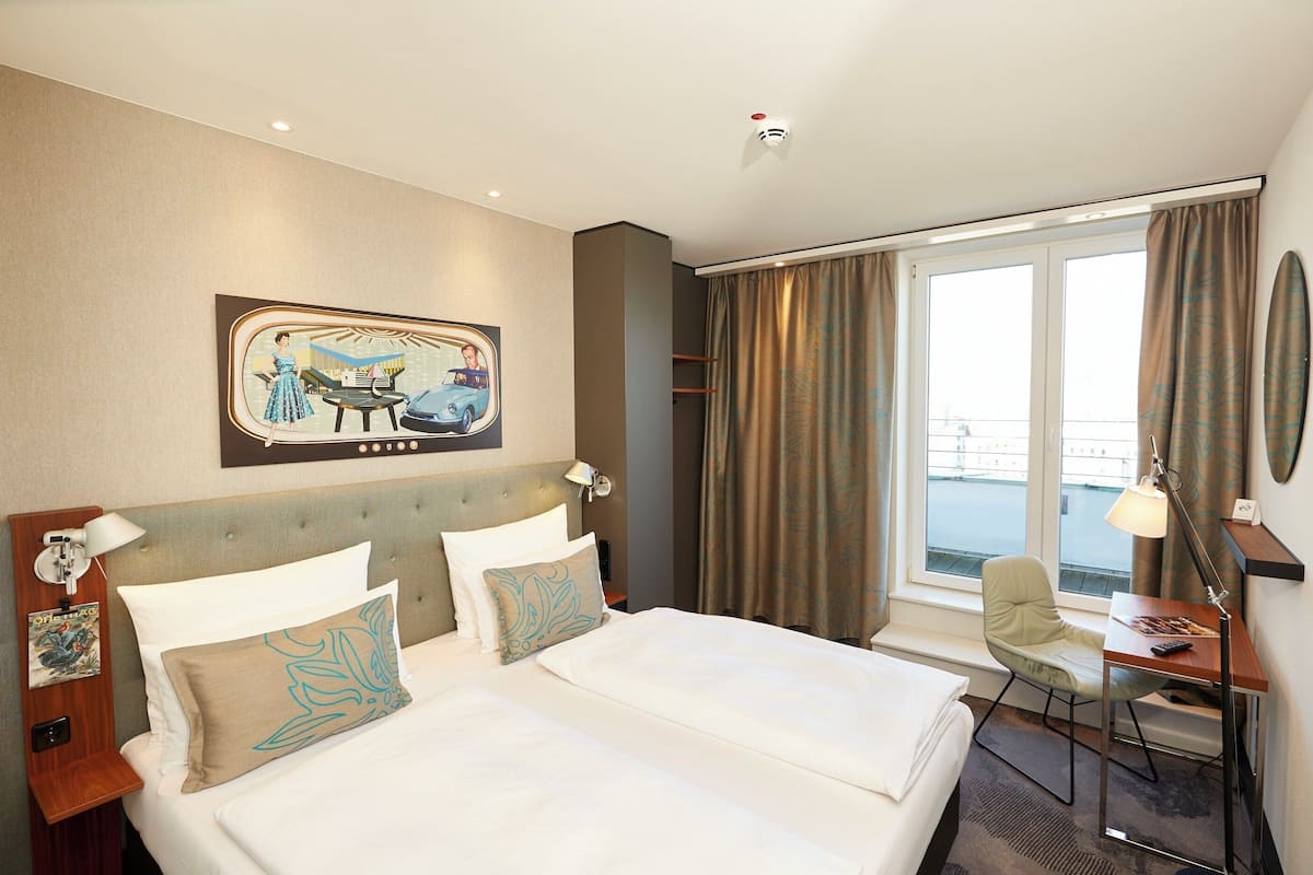 Quarto de hotel com duas camas, uma escrivaninha, uma cadeira, uma janela com cortinas, um quadro na parede e uma mesa de cabeceira com abajur.
