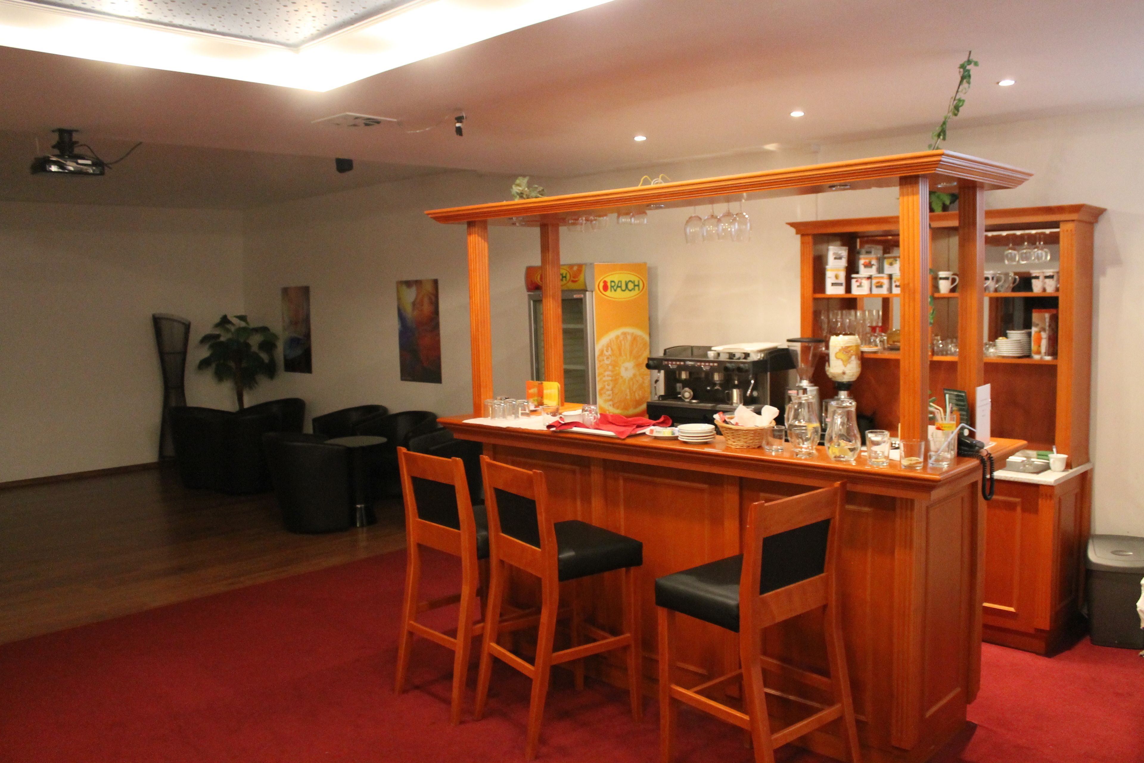 Uma área de bar com banquetas de madeira, um balcão com diversos itens e uma estante com copos.