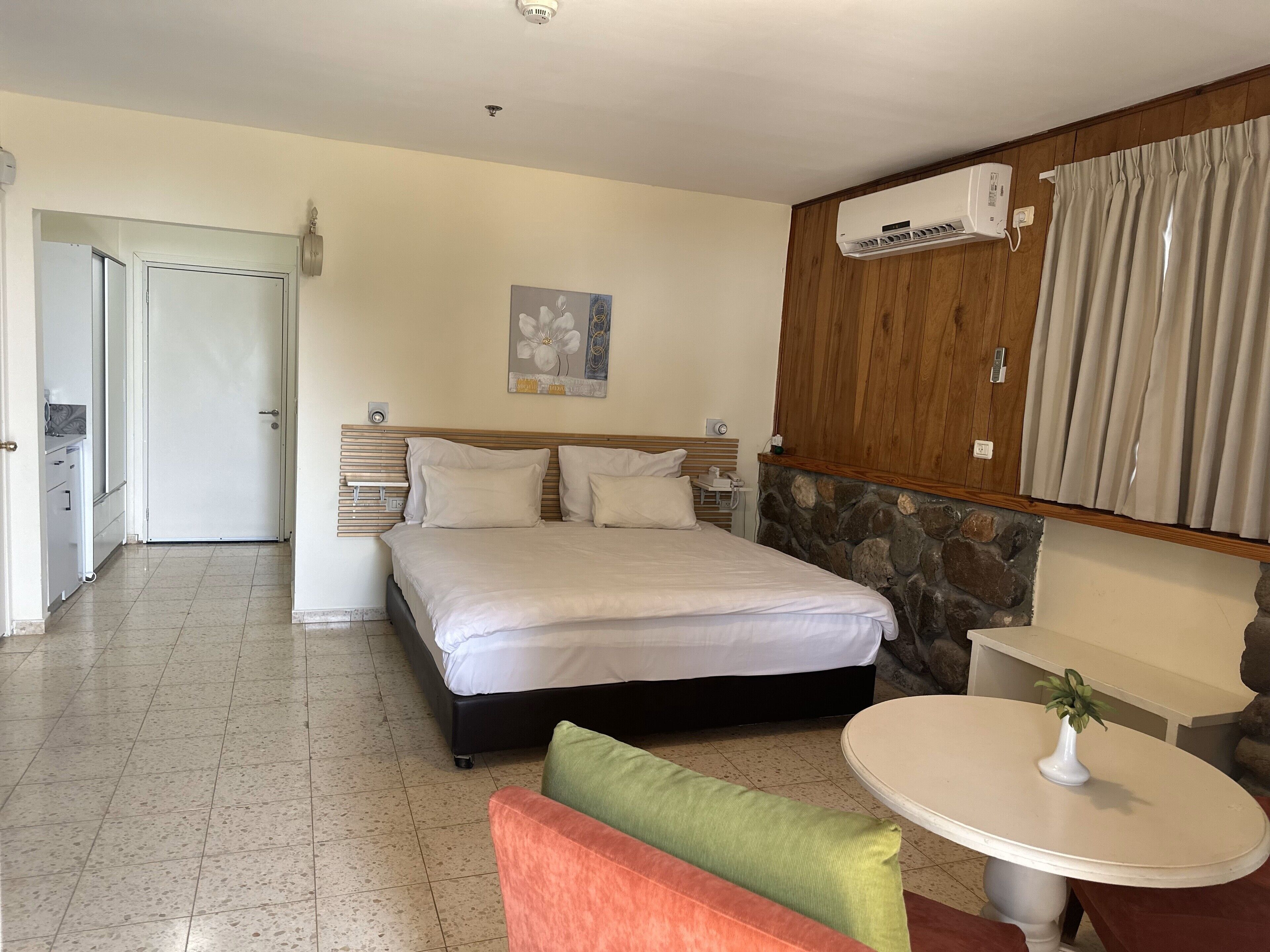 Quarto de hotel com cama, uma mesinha, uma cadeira, um quadro na parede e um ar condicionado.