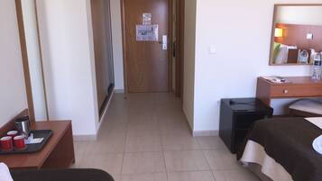 Quarto de hotel com cama, mesa de cabeceira e escrivaninha com cadeira. Há uma porta no final do quarto e um espelho na parede.